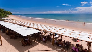 Um restaurante à beira-mar com mesas ao ar livre e guarda-sóis, com vista para uma praia arenosa e o oceano.
