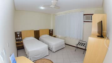 Quarto de hotel com duas camas de solteiro, uma escrivaninha com cadeira, televisão, ventoinha no teto e janela com cortinas.