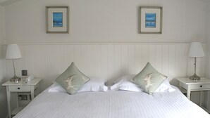 Uma cama de casal com roupa de cama branca, duas mesas de cabeceira com abajures e dois quadros na parede.