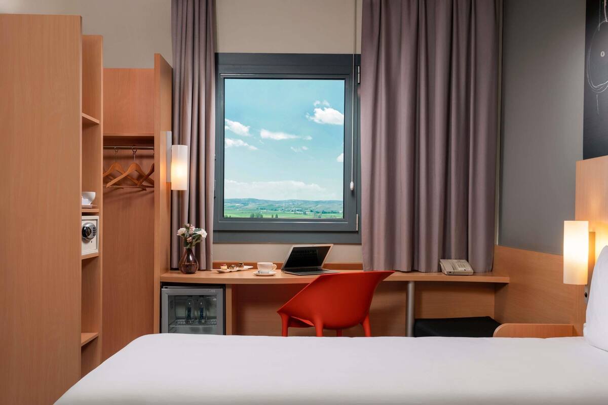Quarto de hotel com cama, escrivaninha com laptop, cadeira, armário e janela com vista para o campo.