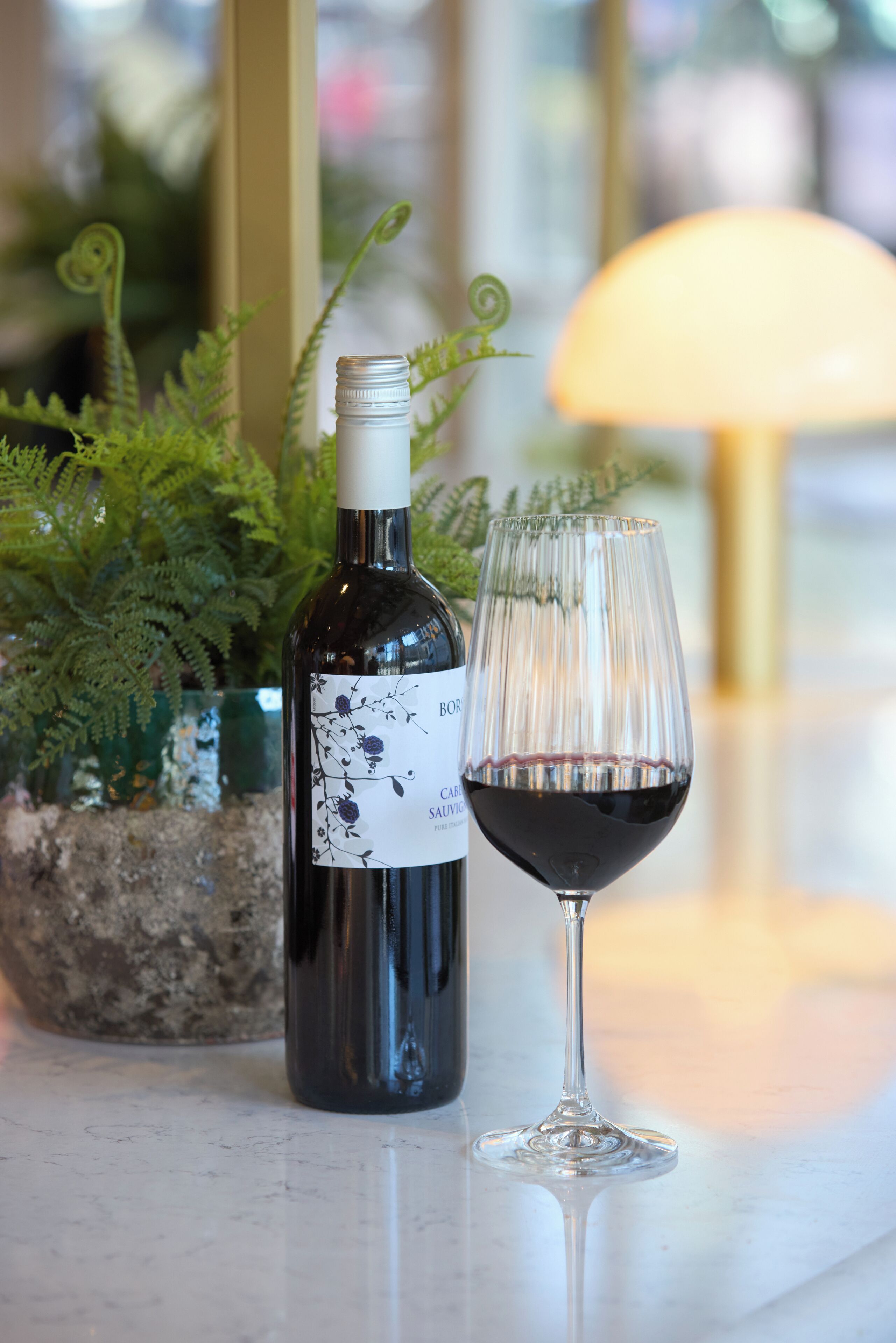 Uma garrafa de vinho e um copo sobre uma mesa, com uma luminária ao fundo.