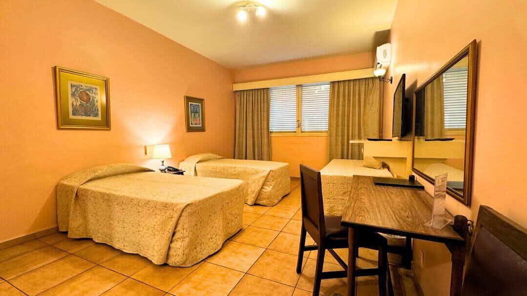 Quarto de hotel com duas camas, uma escrivaninha, uma cadeira, um abajur, um espelho e uma janela com cortinas.