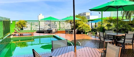 Área de piscina no terraço com guarda-sóis verdes, espreguiçadeiras e mobiliário de madeira.