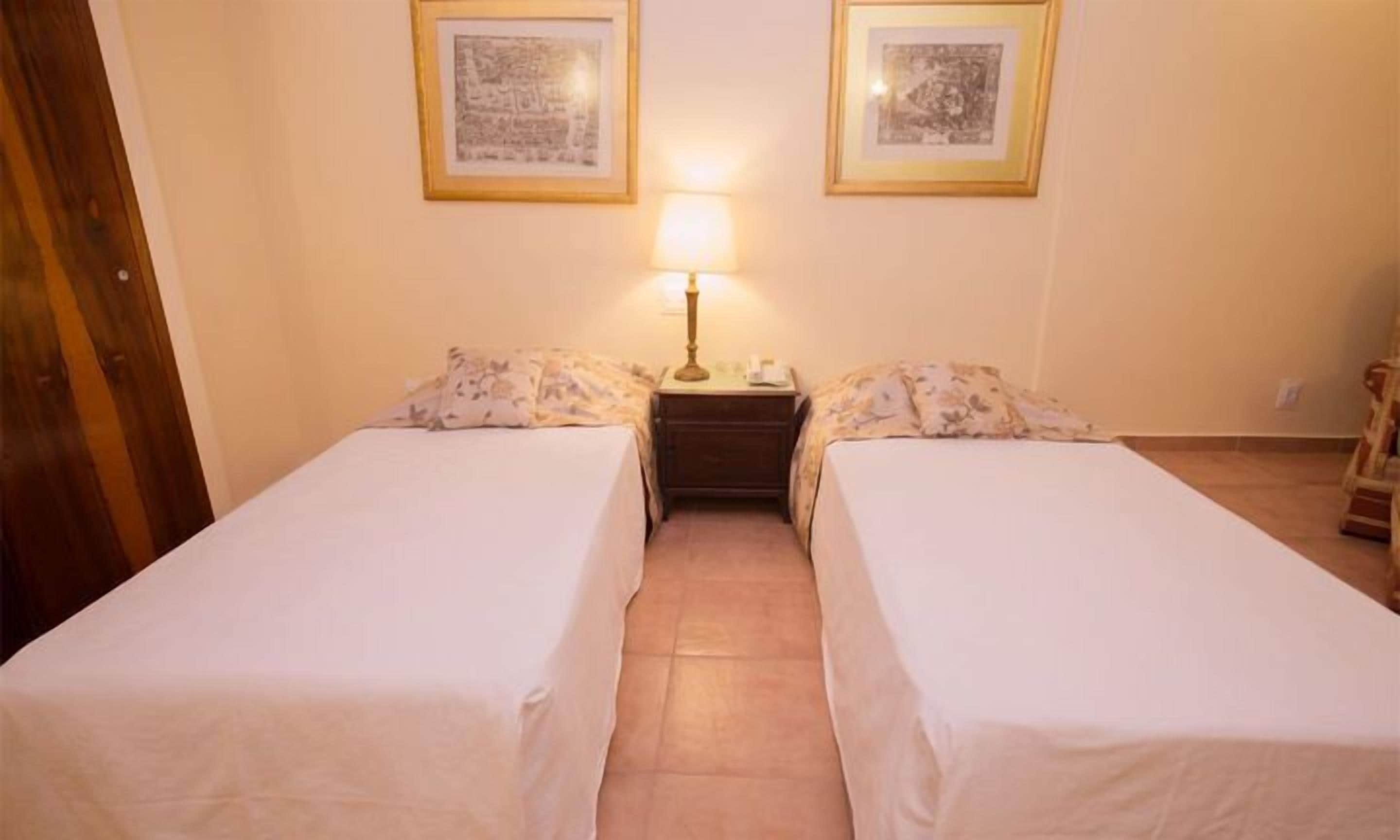 Quarto com duas camas de solteiro, mesa de cabeceira com abajur e duas imagens emolduradas na parede.