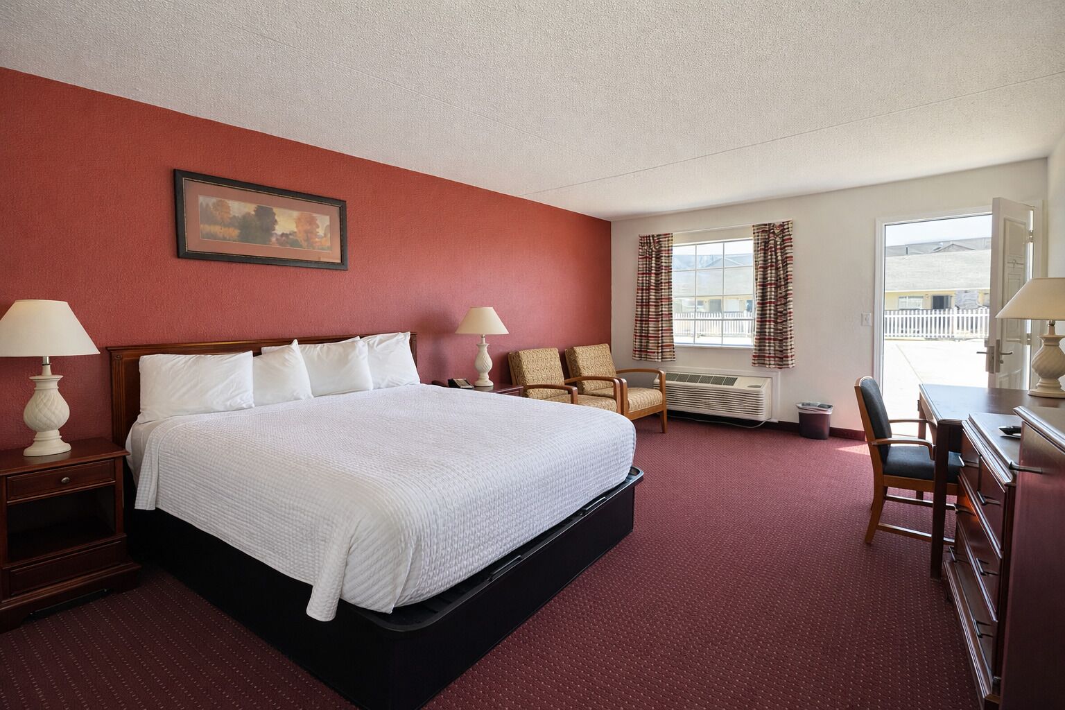 Quarto de hotel com uma cama grande, duas mesas de cabeceira com luminárias, uma escrivaninha com cadeira, uma janela com cortinas e um radiador.