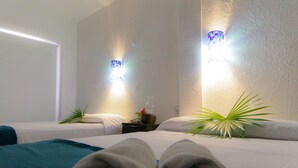 Quarto de hotel com duas camas, uma mesinha com um vaso e uma garrafa, e duas luminárias fixadas na parede.