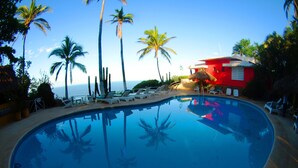 Piscina com espreguiçadeiras e palmeiras, cercada por um edifício vermelho e vista para o oceano.