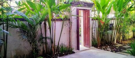 Uma passagem leva a uma pequena construção com porta vermelha, cercada por vegetação exuberante e um chão de azulejos.