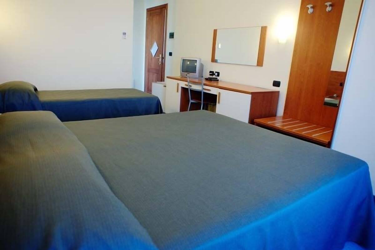 Quarto de hotel com duas camas, uma secretária com computador, um espelho e um guarda-roupa de madeira.