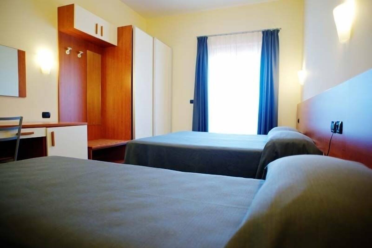 Quarto de hotel com duas camas, uma escrivaninha, um guarda-roupa e uma janela com cortinas.