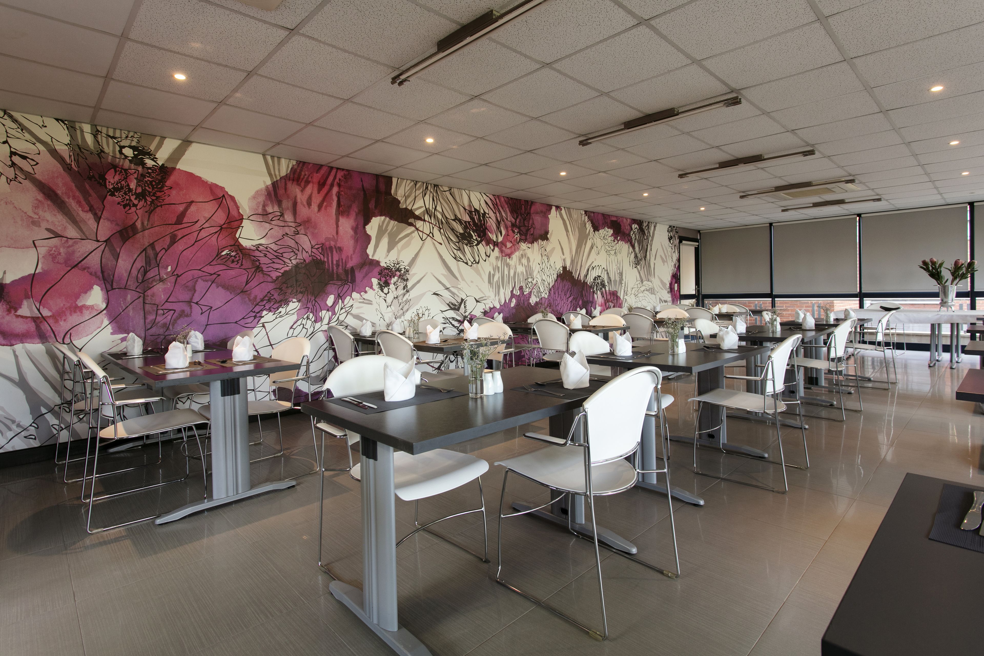 Uma sala de conferência moderna com cadeiras brancas, mesas escuras e um grande mural floral na parede.