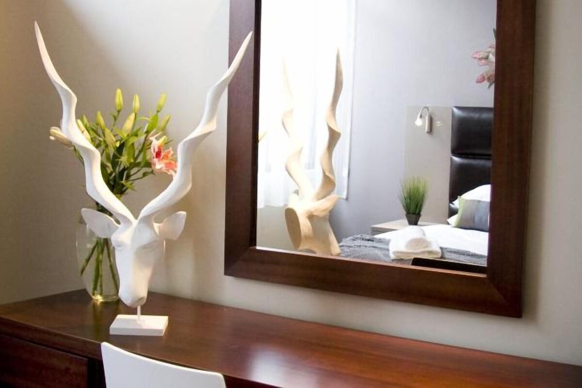 Uma escrivaninha de madeira com um vaso branco cheio de flores e um espelho refletindo uma cama com um travesseiro e uma pequena planta.