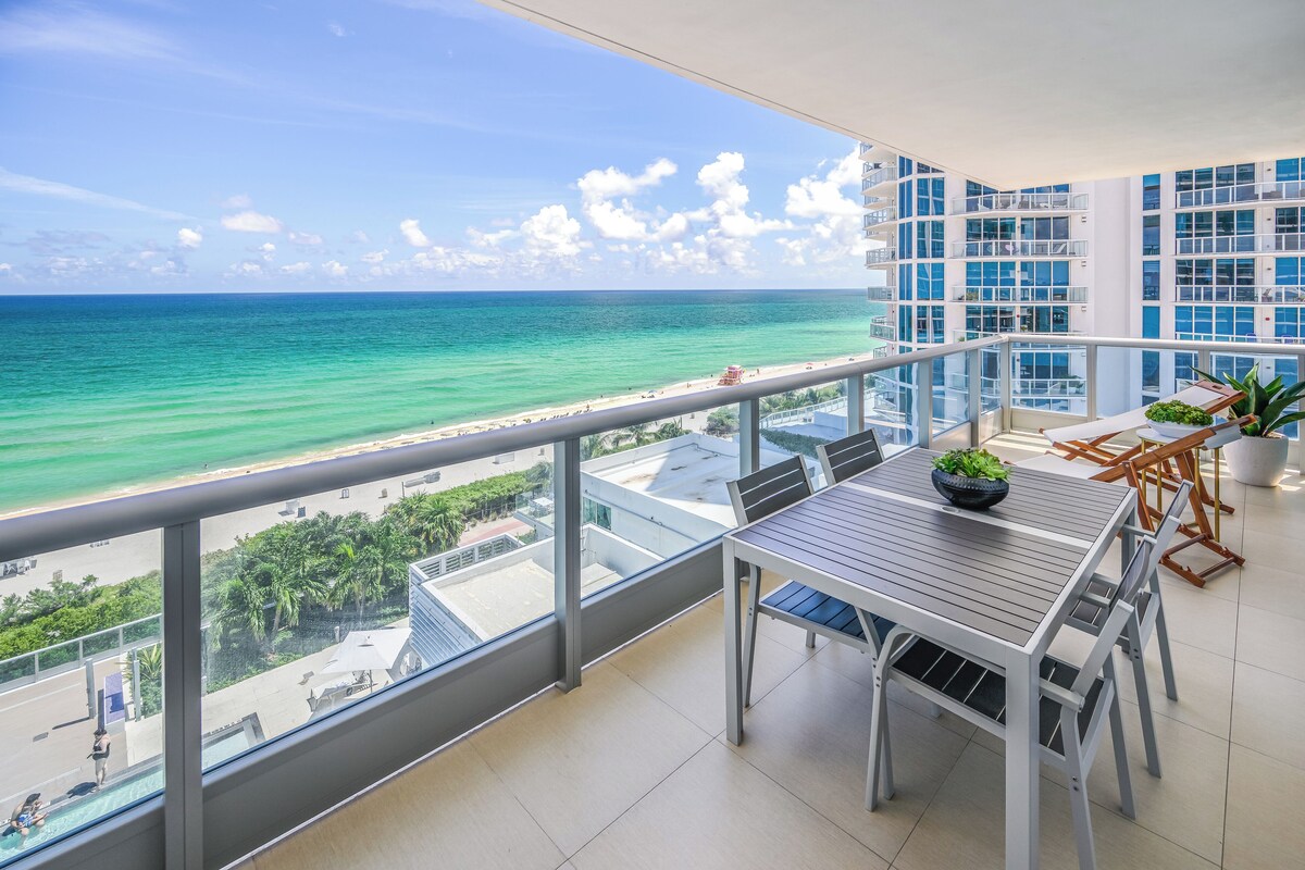 Varanda com vista para a praia e oceano, mesa de jantar com cadeiras e um vaso de planta.