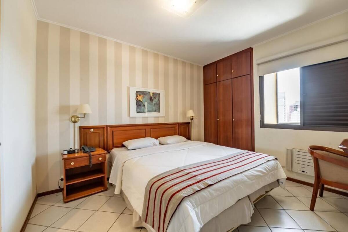 Quarto de hotel com cama, escrivaninha, cadeira, luminária e janela.