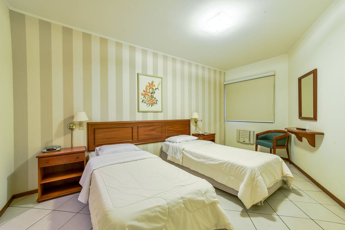 Quarto de hotel com duas camas, cabeceira de madeira, mesa de cabeceira com abajur, escrivaninha com cadeira, janela com cortina e espelho fixado na parede.