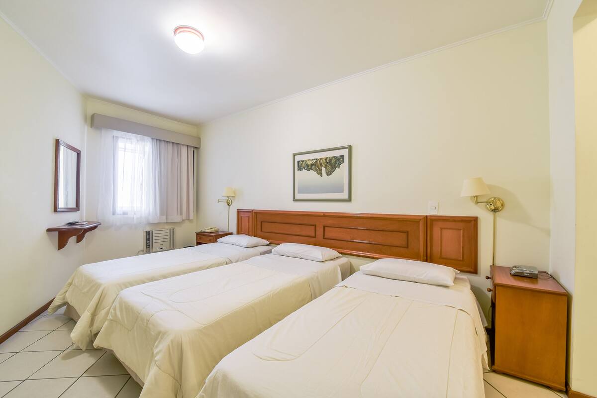 Quarto de hotel com duas camas, cabeceira de madeira, mesa de cabeceira com luminária, janela com cortinas, um quadro na parede e uma pequena estante.