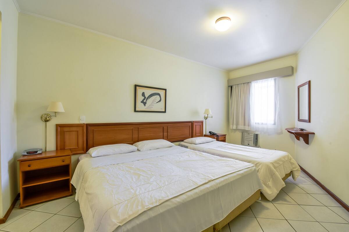 Quarto de hotel com duas camas, cabeceira de madeira, mesa de cabeceira com abajur, prateleira pequena e janela com cortinas.