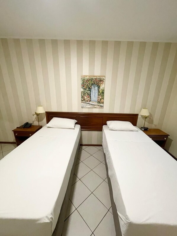 Quarto de hotel com duas camas de solteiro, papel de parede listrado, uma pequena janela com vista para um portão e mesas de cabeceira com luminárias.