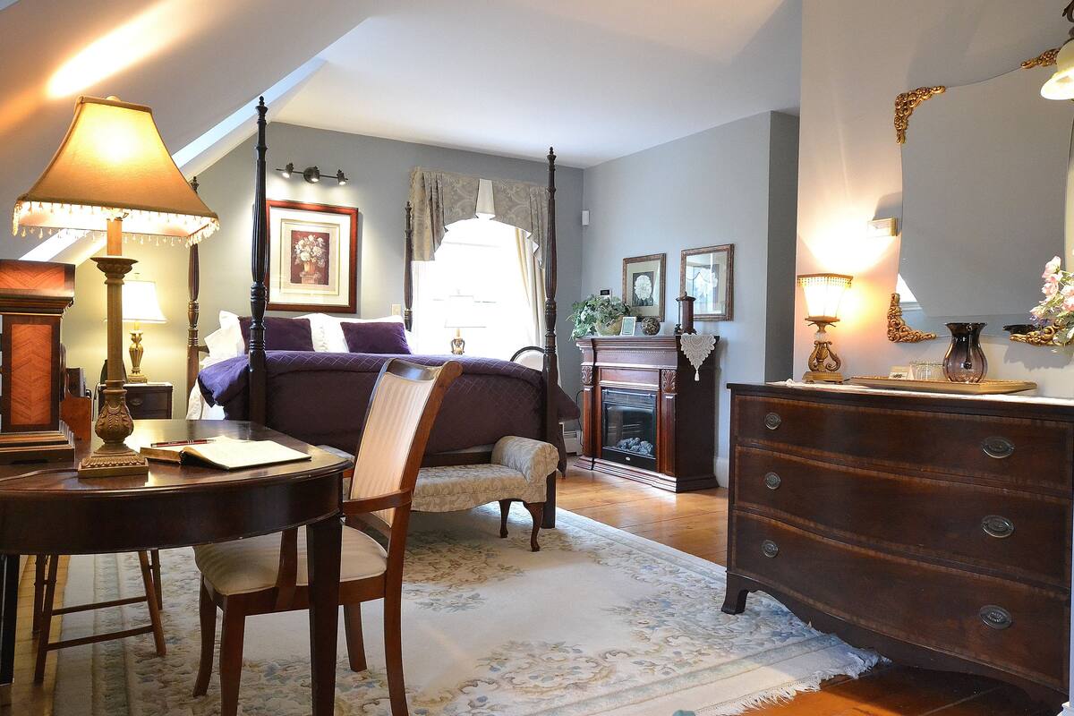 Um quarto com uma cama de dossel, uma cômoda de madeira, uma escrivaninha com abajur e uma lareira.