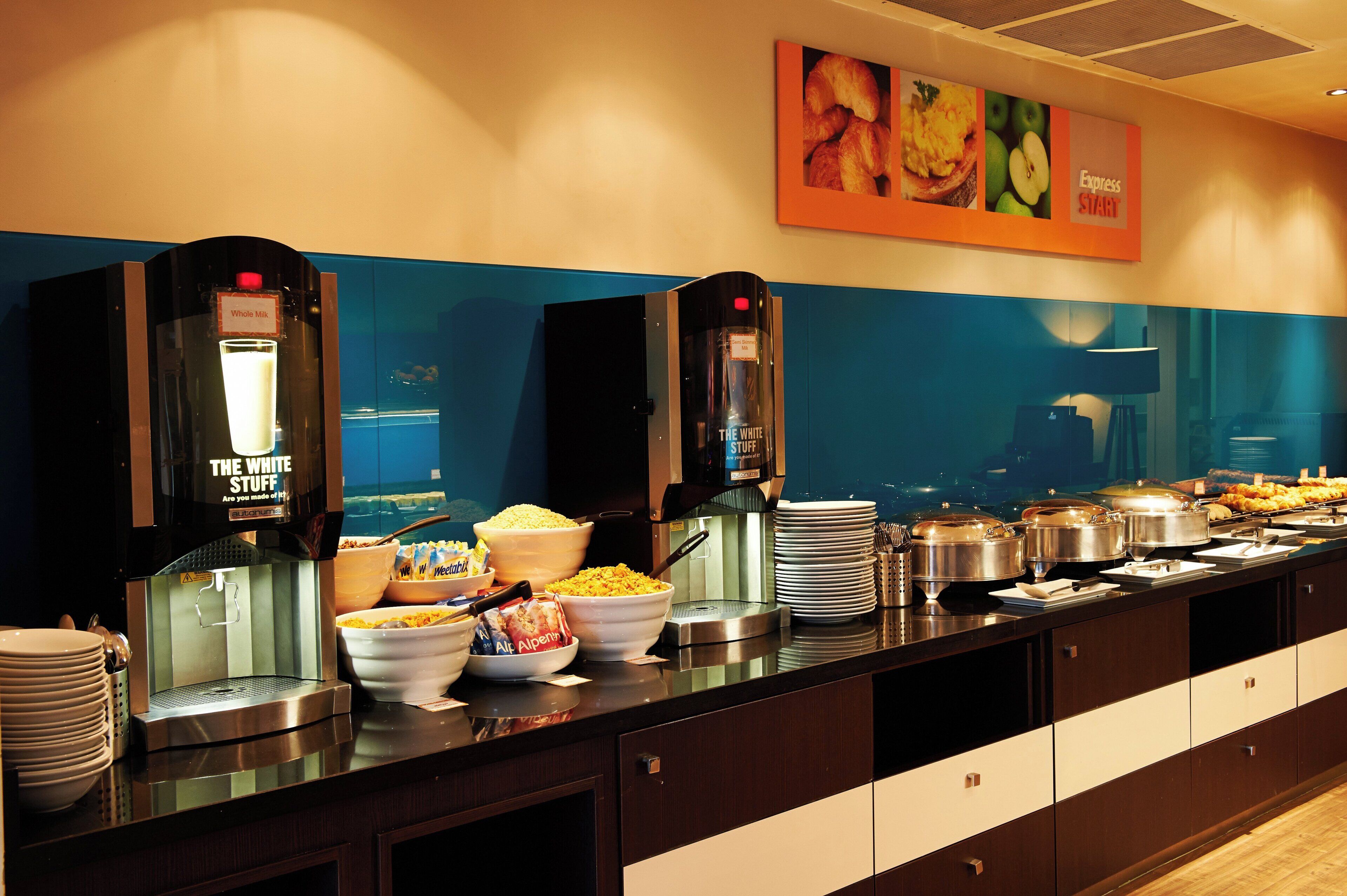 Disposição de buffet com diversas estações de comida, incluindo um dispensador de leite integral e uma máquina de café self-service.