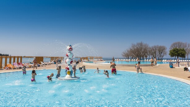 Piscina com uma fonte de água, rodeada por espreguiçadeiras e uma área de praia arenosa.
