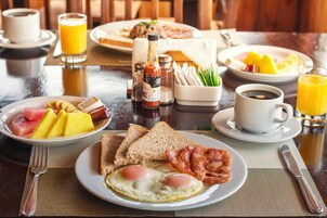 Uma mesa de café da manhã com pratos de ovos, torradas, bacon e frutas. Há também acompanhamentos, uma xícara de café e um copo de suco de laranja.