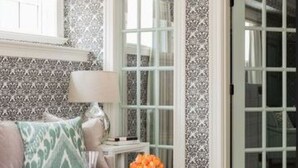 Um quarto com papel de parede estampado, um banco de couro marrom, uma mesa branca com uma bandeja de comida e uma janela com cortinas.