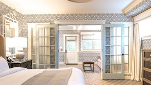 Quarto de hotel com uma cama grande, uma escrivaninha com luminária, uma cadeira e um guarda-roupa. Há uma porta que dá para o banheiro e uma janela com cortinas.