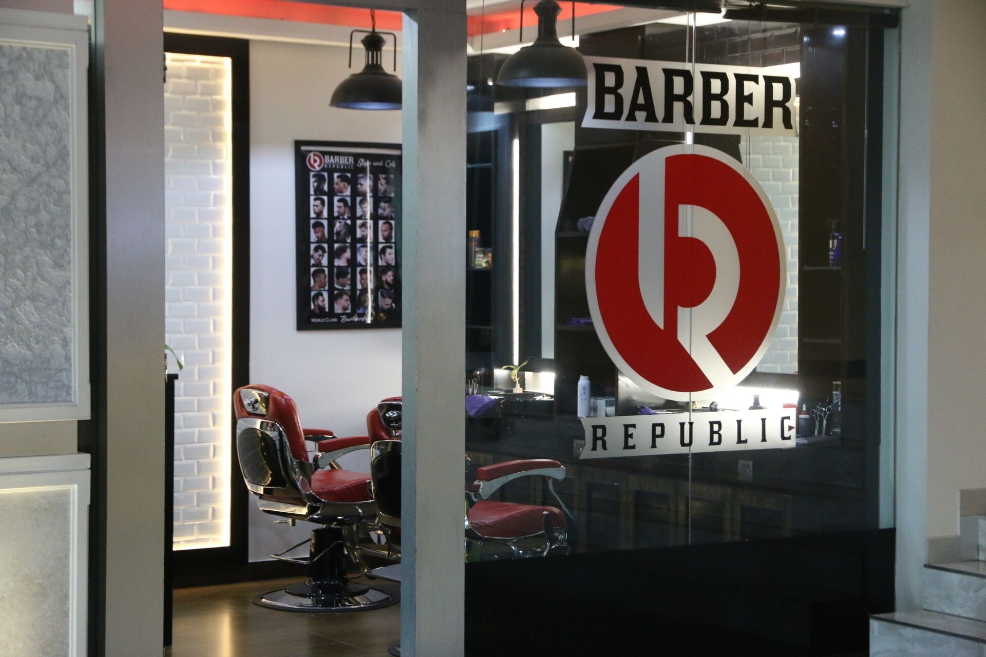 Interior de barbearia com um grande logotipo de barbeiro vermelho e branco, duas cadeiras de barbeiro e um pôster emoldurado na parede.