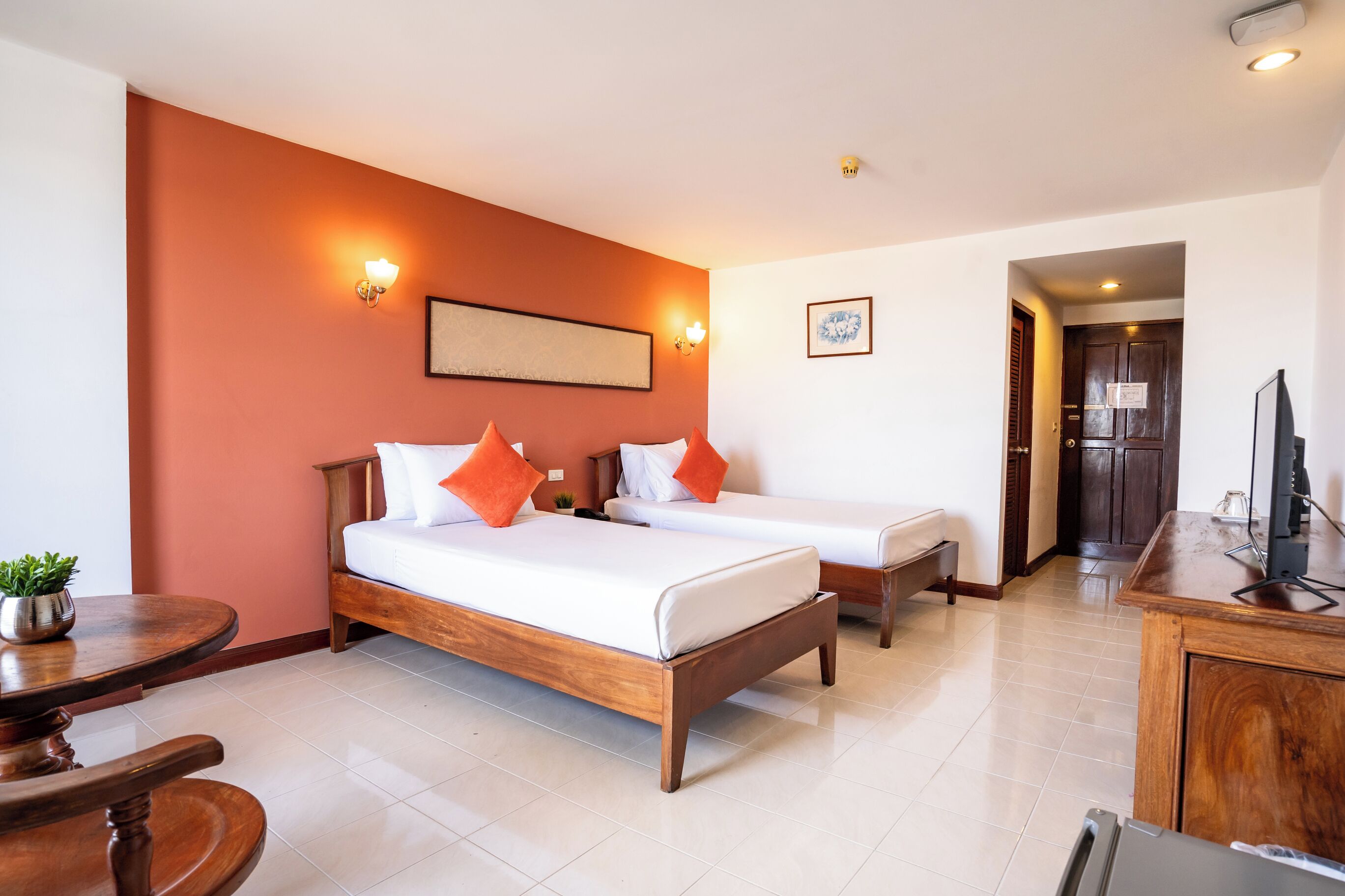 Quarto de hotel com duas camas, uma escrivaninha de madeira, uma televisão e uma pequena mesa com uma planta.
