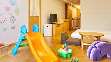 Quarto de hotel com cama, escrivaninha, televisão, cadeira, mesa, sofá e área de recreação infantil com brinquedos, além de parede decorada com desenhos de crianças.