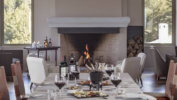 Uma sala de jantar com lareira, cadeiras de madeira, uma mesa comprida posta para refeição e garrafas de vinho.