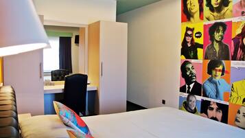 Quarto de hotel com uma cama, uma mesa e uma cadeira. Há cartazes na parede.