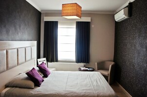 Quarto de hotel com uma cama grande, uma mesa de cabeceira, uma cadeira, janela com cortinas e uma luminária no teto.