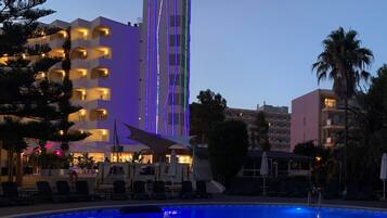 Um hotel com piscina, área de estar externa e um edifício alto com linhas verticais iluminadas.