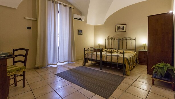 Um quarto com uma cama, uma escrivaninha, uma cadeira, mesa de cabeceira, abajur, planta e uma janela com cortinas.