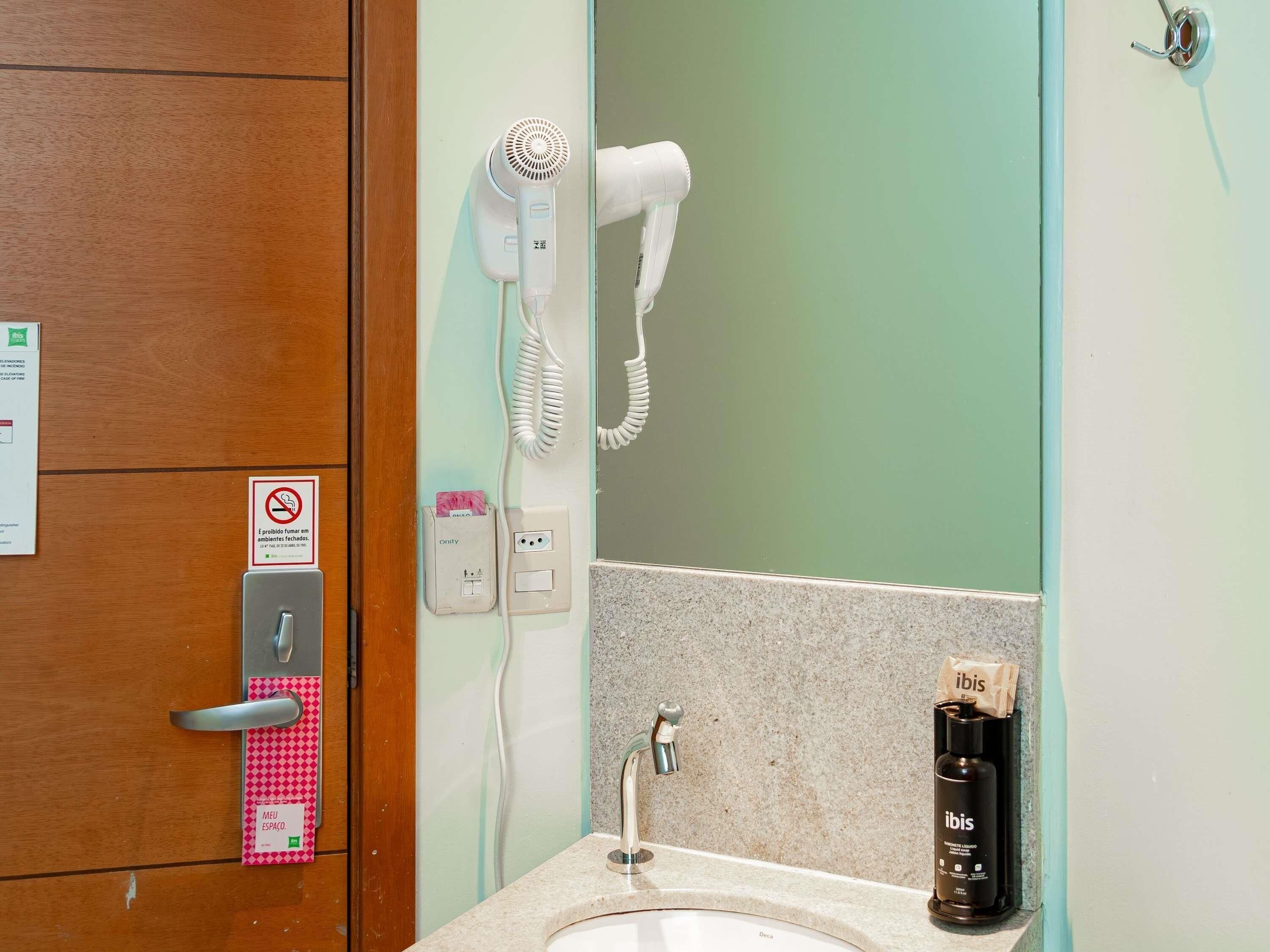Uma casa de banho com secador de cabelo fixado na parede, uma pia com dispensador de sabão e uma porta com puxador.