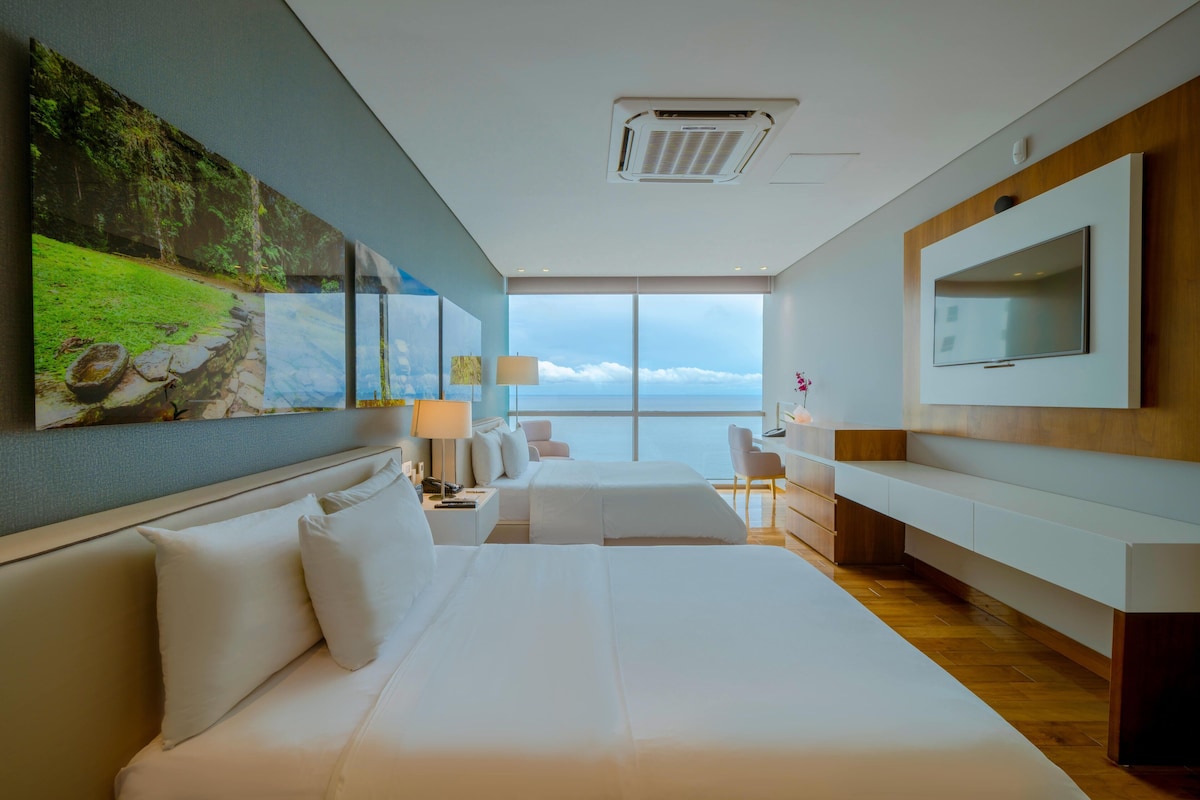 Quarto de hotel moderno com uma cama grande, escrivaninha, cadeira, televisão e uma janela ampla com vista para o céu.