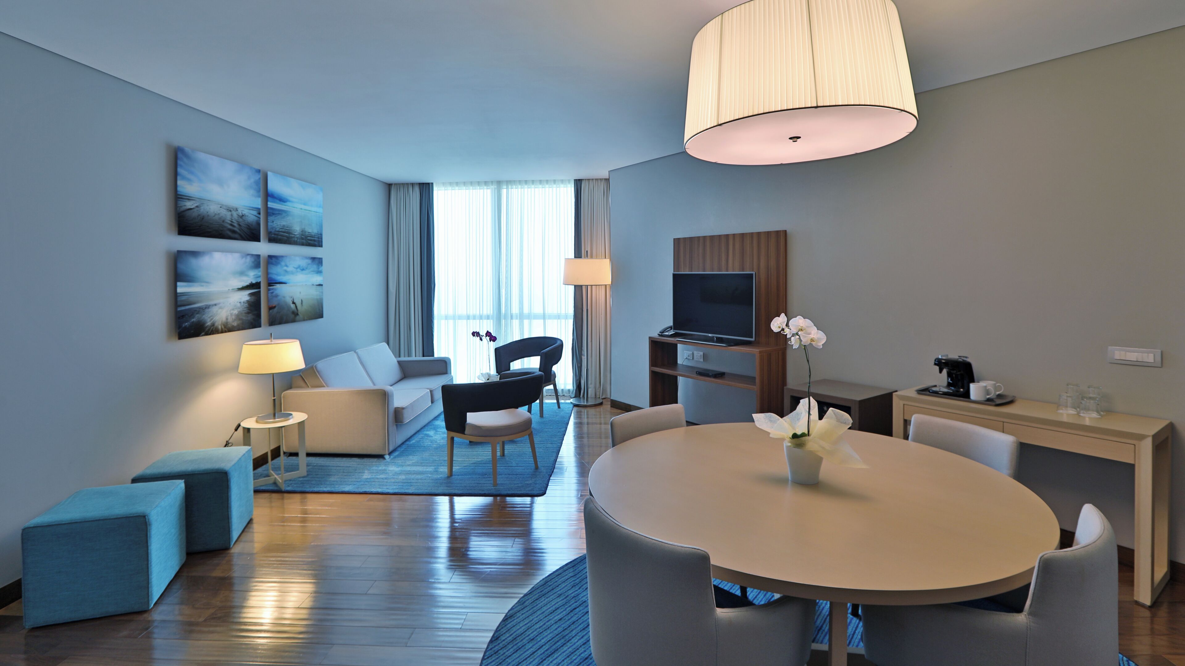 Quarto de hotel moderno com sofá, mesa de jantar, televisão e uma janela grande.
