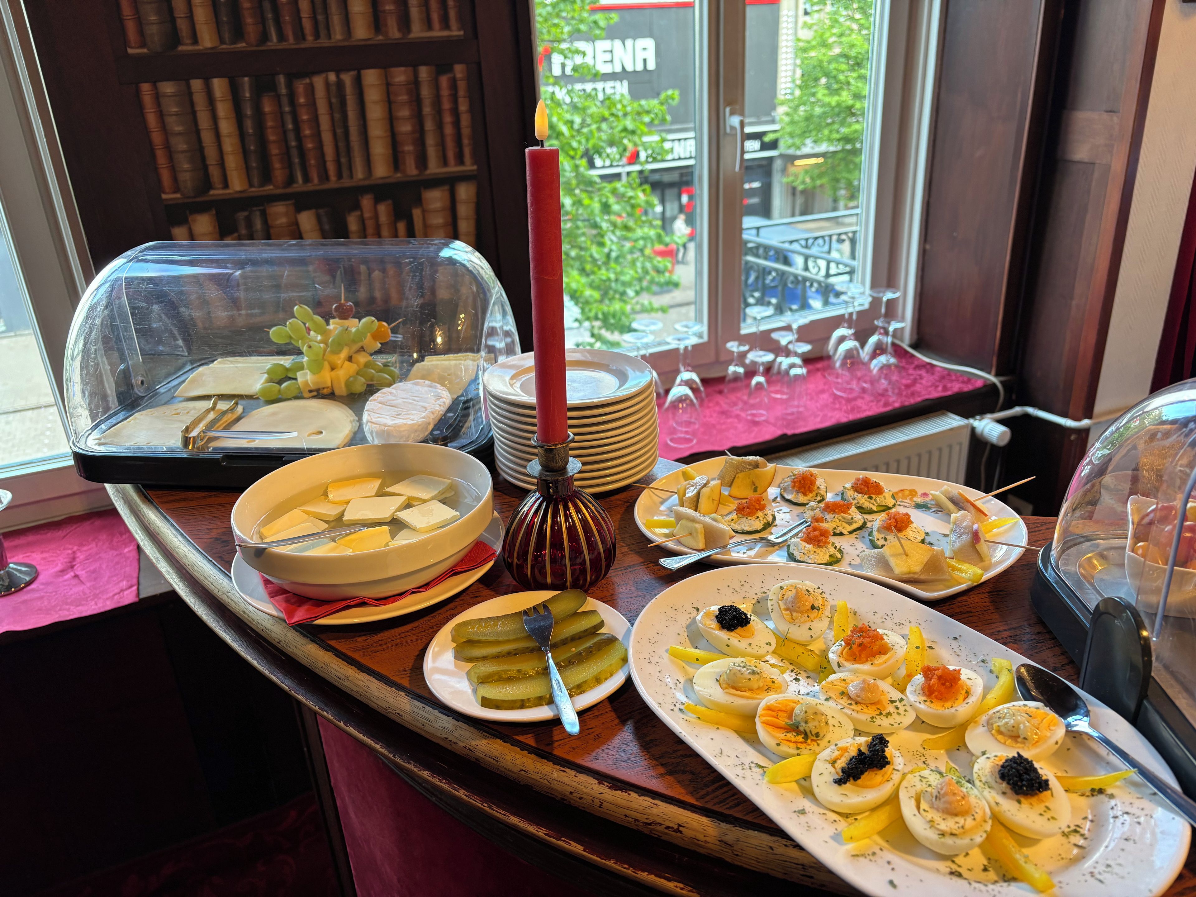 Mesa de buffet com diversos alimentos, incluindo queijo, picles e ovos recheados, em um ambiente com estante de livros e vista para o exterior.