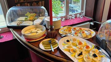 Mesa de buffet com diversos alimentos, incluindo queijo, picles e ovos recheados, em um ambiente com estante de livros e vista para o exterior.