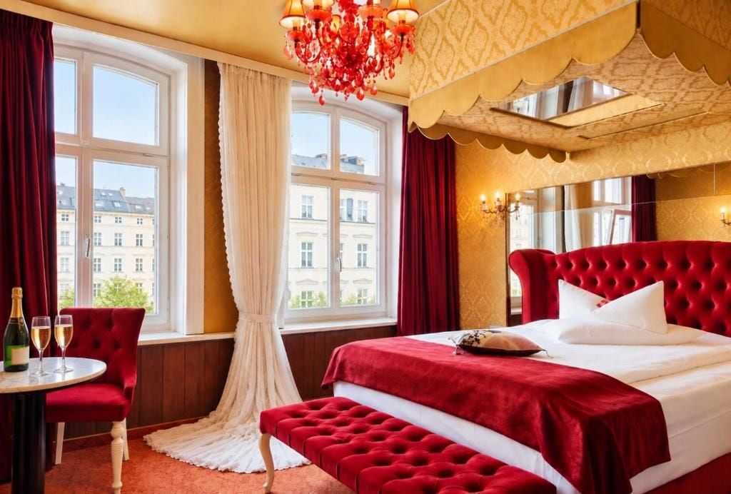 Quarto de hotel com uma cama grande, um banquinho estofado em vermelho, uma mesa redonda com garrafa e copos, e vista para prédios por grandes janelas.