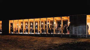 Um edifício moderno com vários espaços com paredes de vidro, cada um iluminado por luminárias pendentes, localizado em um campo gramado à noite.