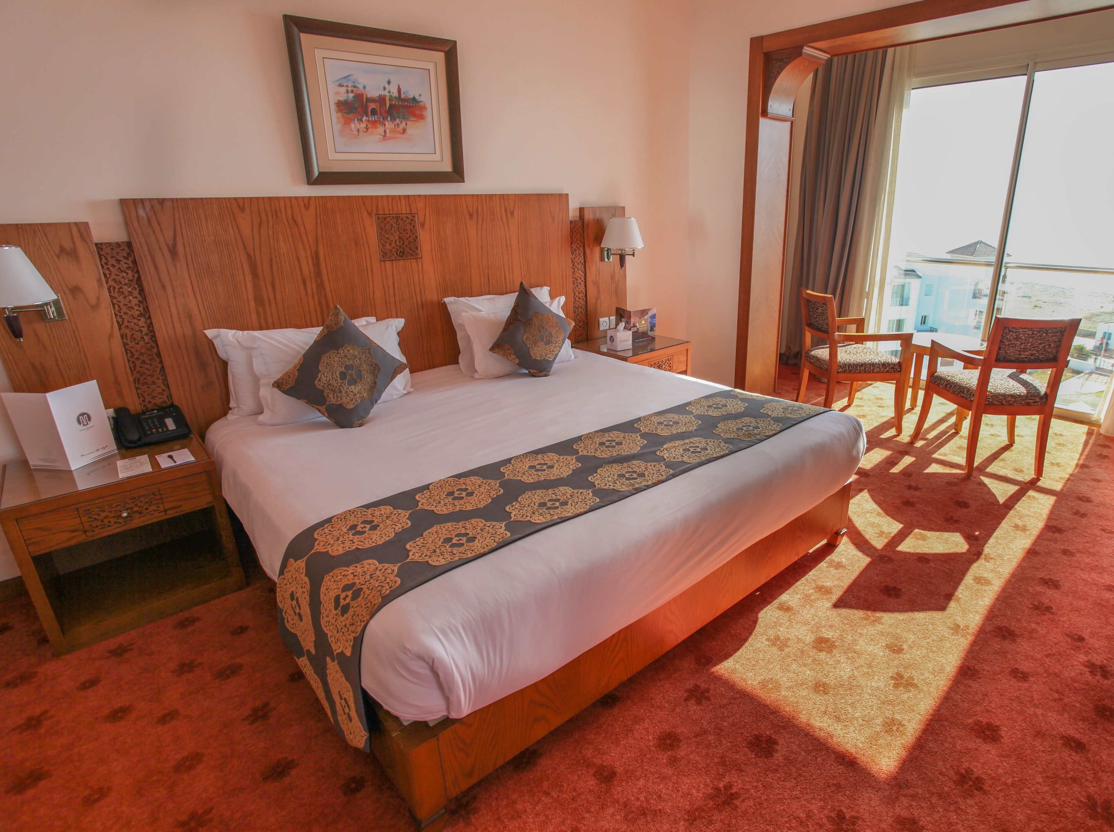 Quarto de hotel com uma cama grande, cabeceira de madeira, duas mesas de cabeceira, um criado-mudo, uma escrivaninha, uma cadeira, uma janela com vista e um quadro na parede.