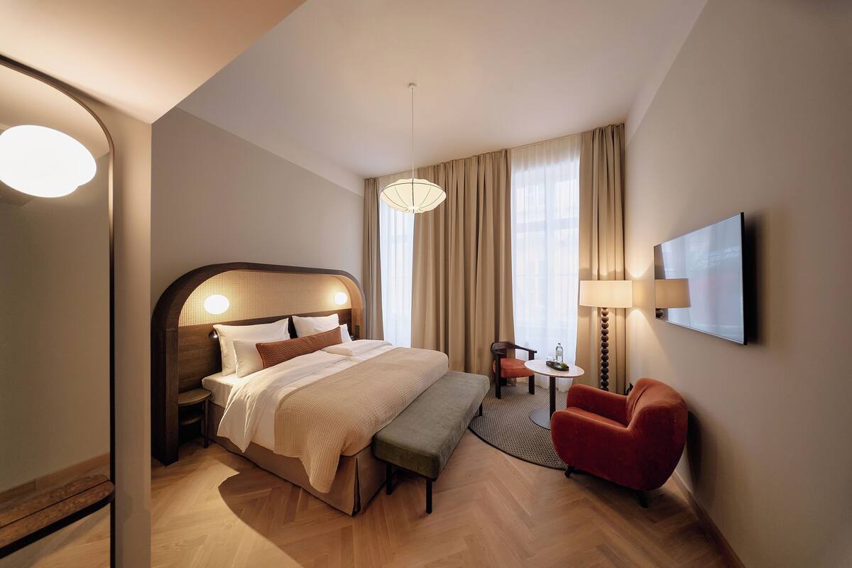Quarto de hotel com cama, mesa de cabeceira, luminária, cadeira e televisão na parede.