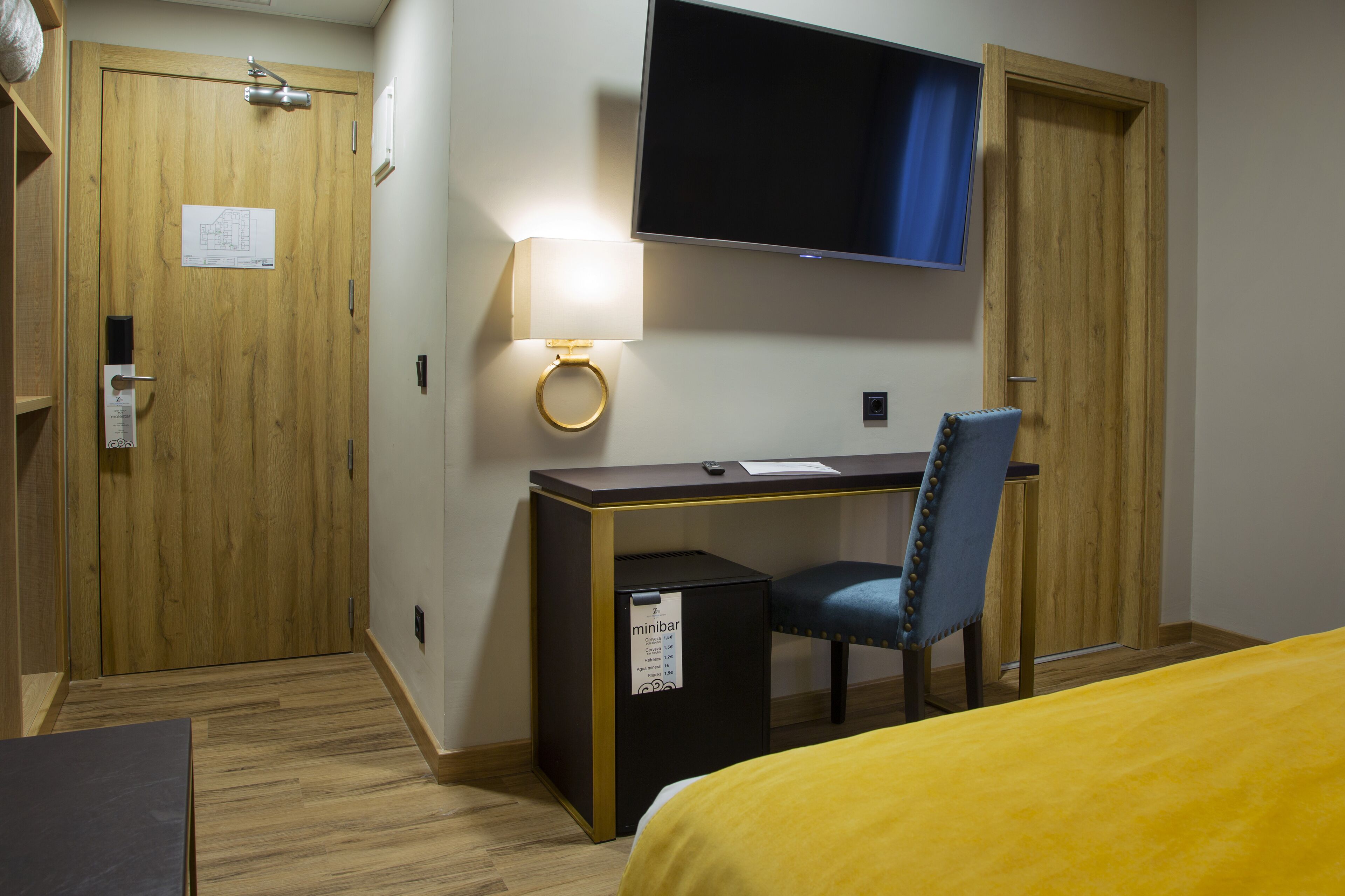 Quarto de hotel com escrivaninha de madeira, uma cadeira, cama com colcha amarela, televisão fixada na parede e minibar.