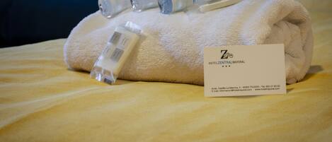 Toalha dobrada sobre a cama, com artigos de higiene pessoal. Cartão de visita do HOTELZENTRALMAYORAL visível.