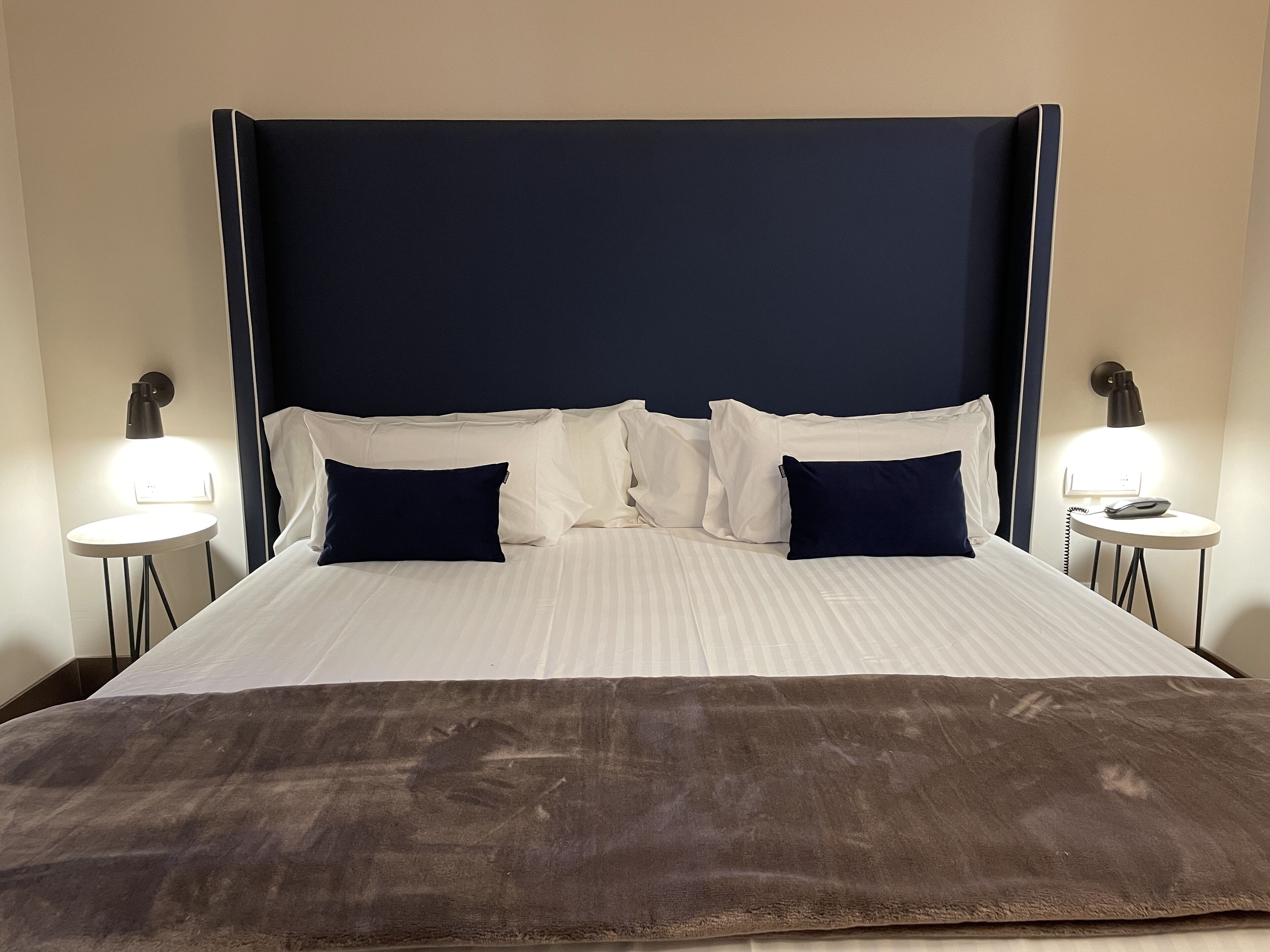 Cama bem arrumada com lençóis brancos e almofadas azul-escuras, acompanhada por duas mesas de cabeceira com abajures, num quarto com cabeceira azul-escura.
