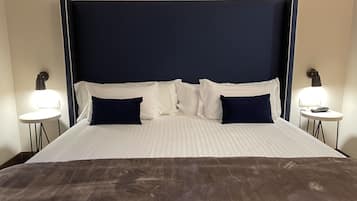 Cama bem arrumada com lençóis brancos e almofadas azul-escuras, acompanhada por duas mesas de cabeceira com abajures, num quarto com cabeceira azul-escura.