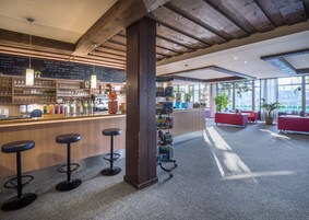 Uma área de bar moderna com banquetas, um balcão com cardápio em lousa e uma sala de estar com sofás vermelhos.
